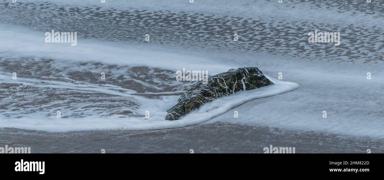 A panoramic image of incoming tide flowing around a rock on a beach ...