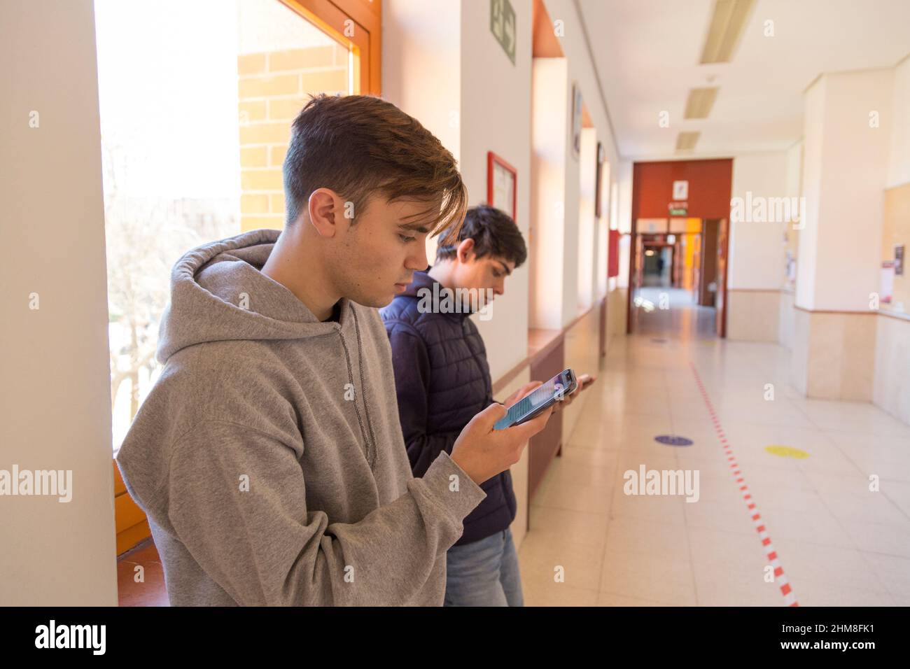 two university student friends leaning against the wall using mobile ...