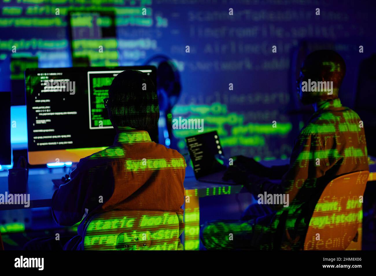 Rear view of two young intercultural programmers sitting in front of computers by desks and ...