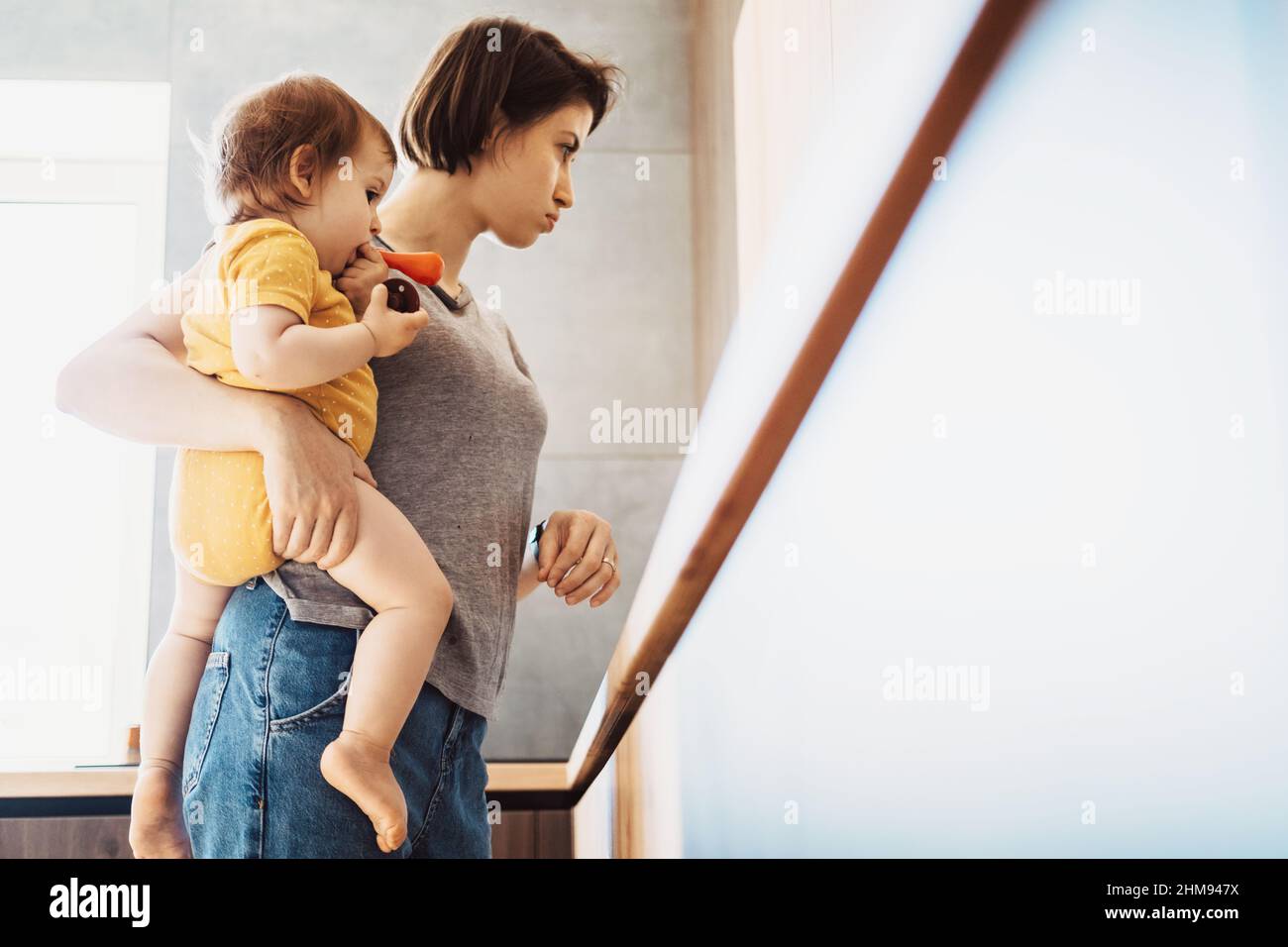 Multitasking mum holding her young baby while standing at the kitchen ...