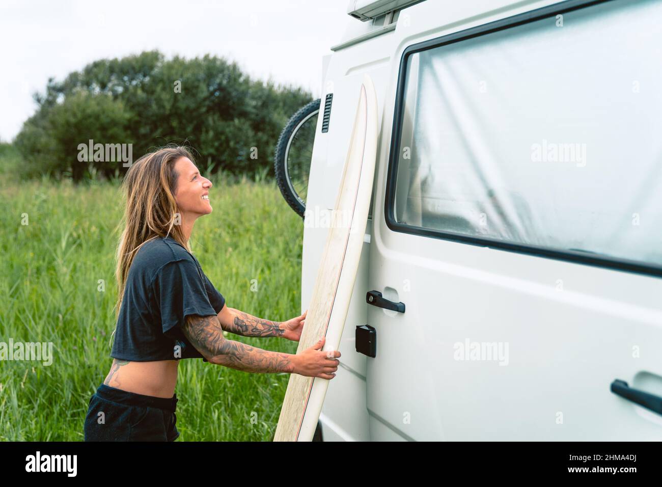 Positive female surfer standing with surfboard near trailer in nature ...