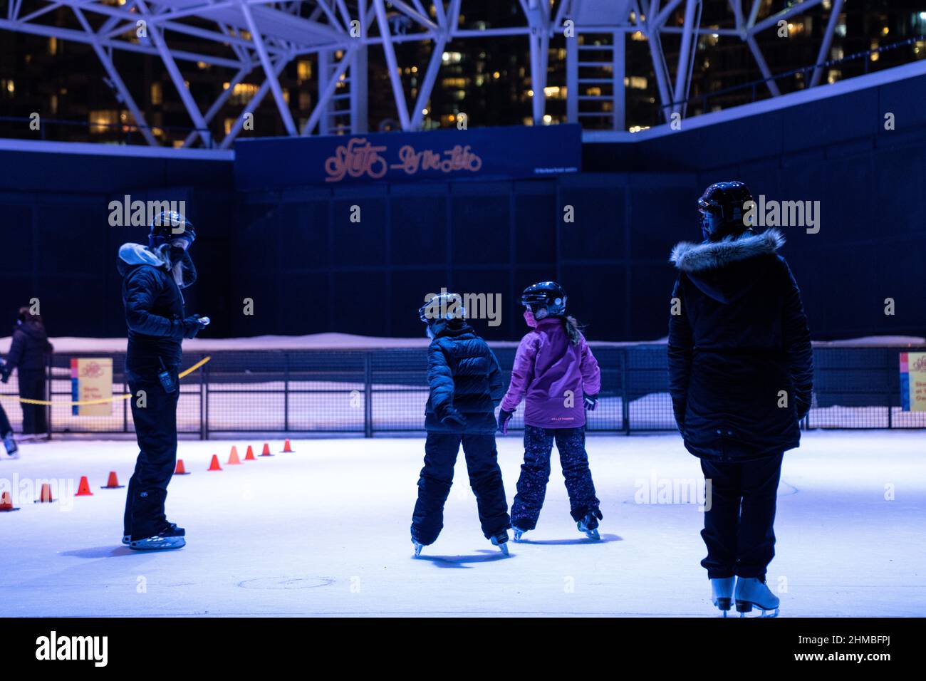 Kids take a skating lesson along a brightly-lit outdoor ice rink.The ...