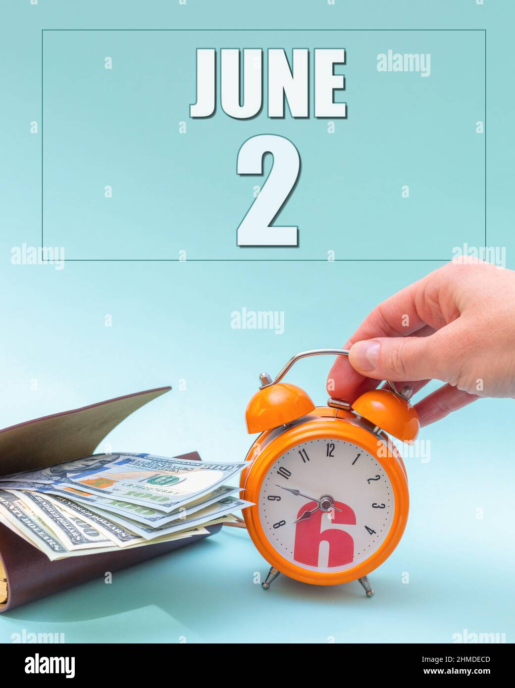 June 2nd. Hand holding an orange alarm clock, a wallet with cash and a ...