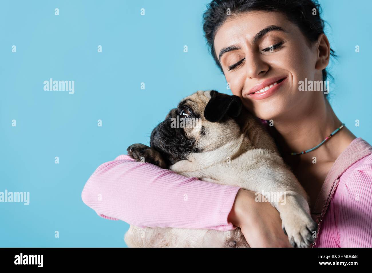happy brunette woman with closed eyes hugging pug dog isolated on blue ...