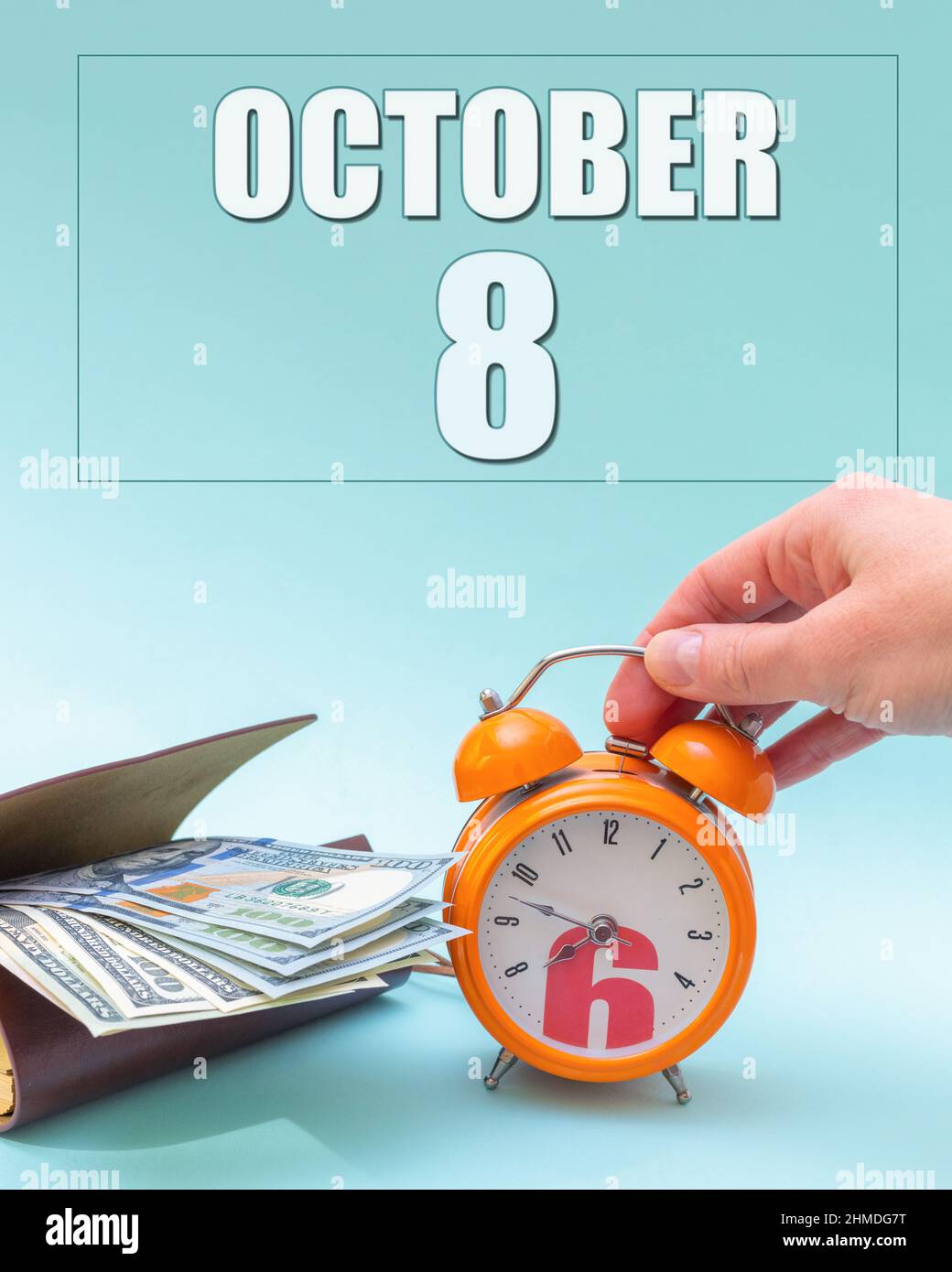 October 8th. Hand holding an orange alarm clock, a wallet with cash and ...