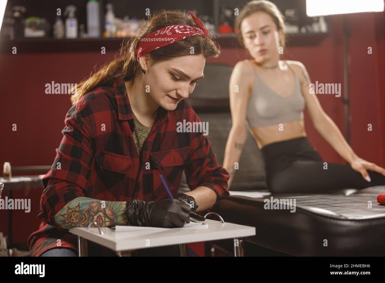 Young lady in top sitting in tattoo salon and watching tattoo artist ...