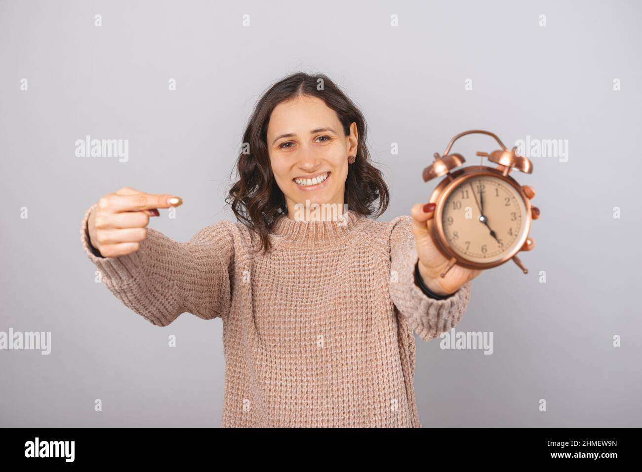 Smiling woman is pointing at the alarm clock she is holding. It is five ...