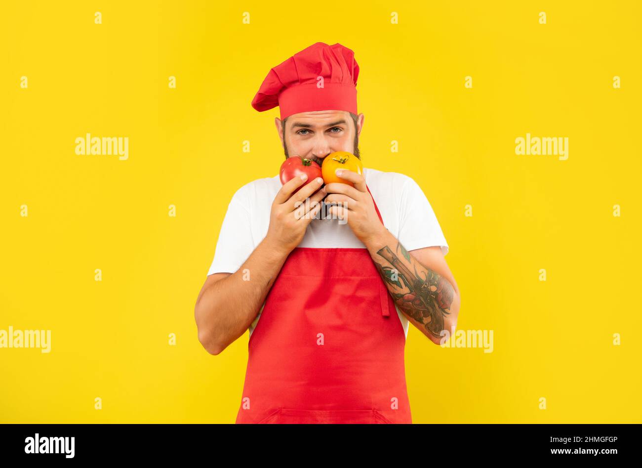 Serious guy in cooking apron and toque smelling tomatoes yellow ...