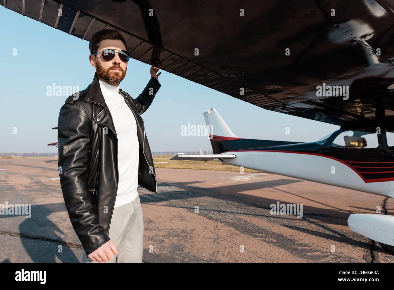bearded pilot in sunglasses and leather jacket standing under aircraft ...
