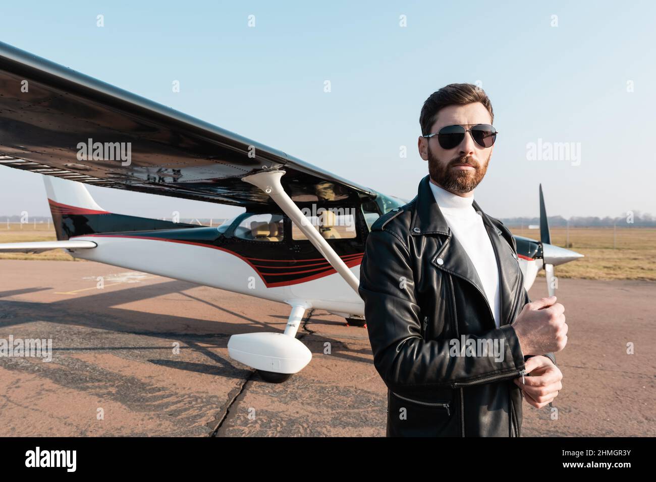 bearded pilot in leather jacket and sunglasses standing near aircraft ...