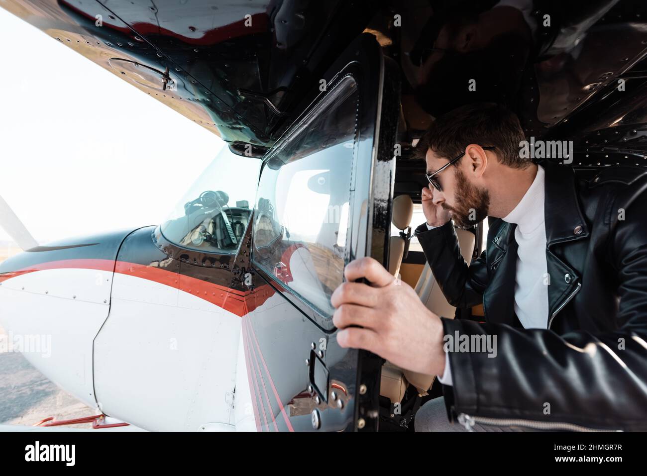 bearded pilot in stylish sunglasses and leather jacket opening door in ...