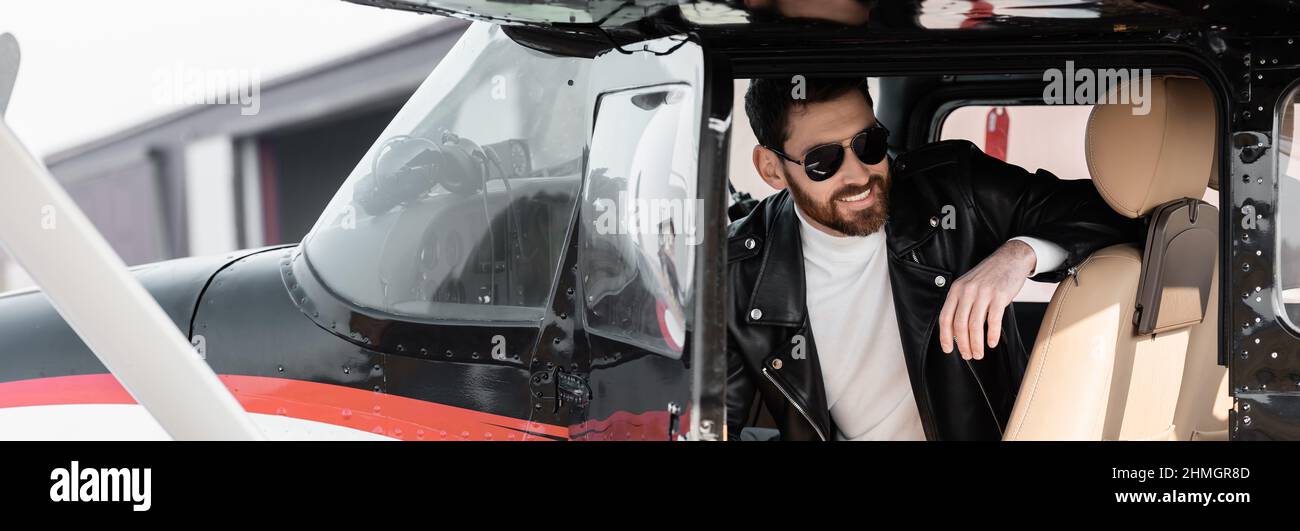 bearded pilot in stylish sunglasses and leather jacket smiling while ...