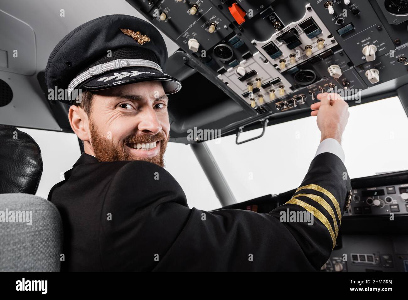 cheerful pilot in cap reaching overhead panel with set of switches in ...