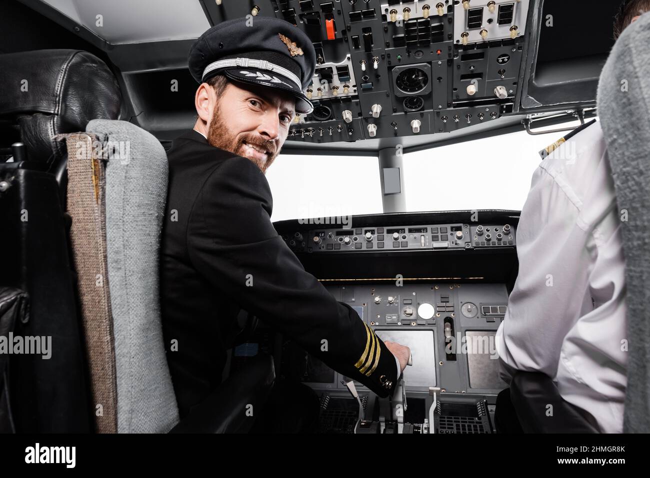 bearded pilot in cap smiling and looking at camera near co-pilot in ...