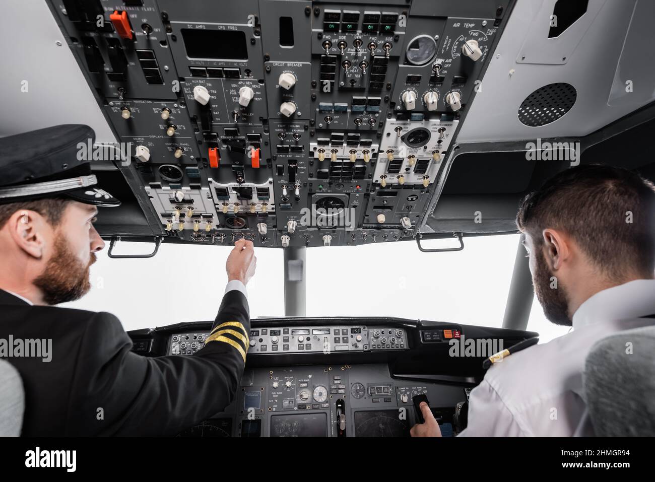 bearded pilot in cap reaching overhead panel near co-pilot in airplane ...