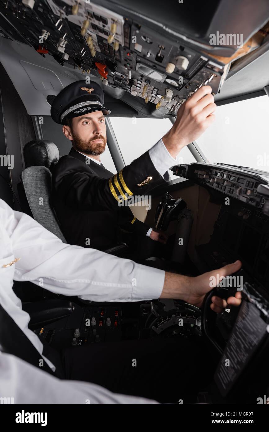 pilot in cap and uniform reaching overhead panel near co-pilot using ...
