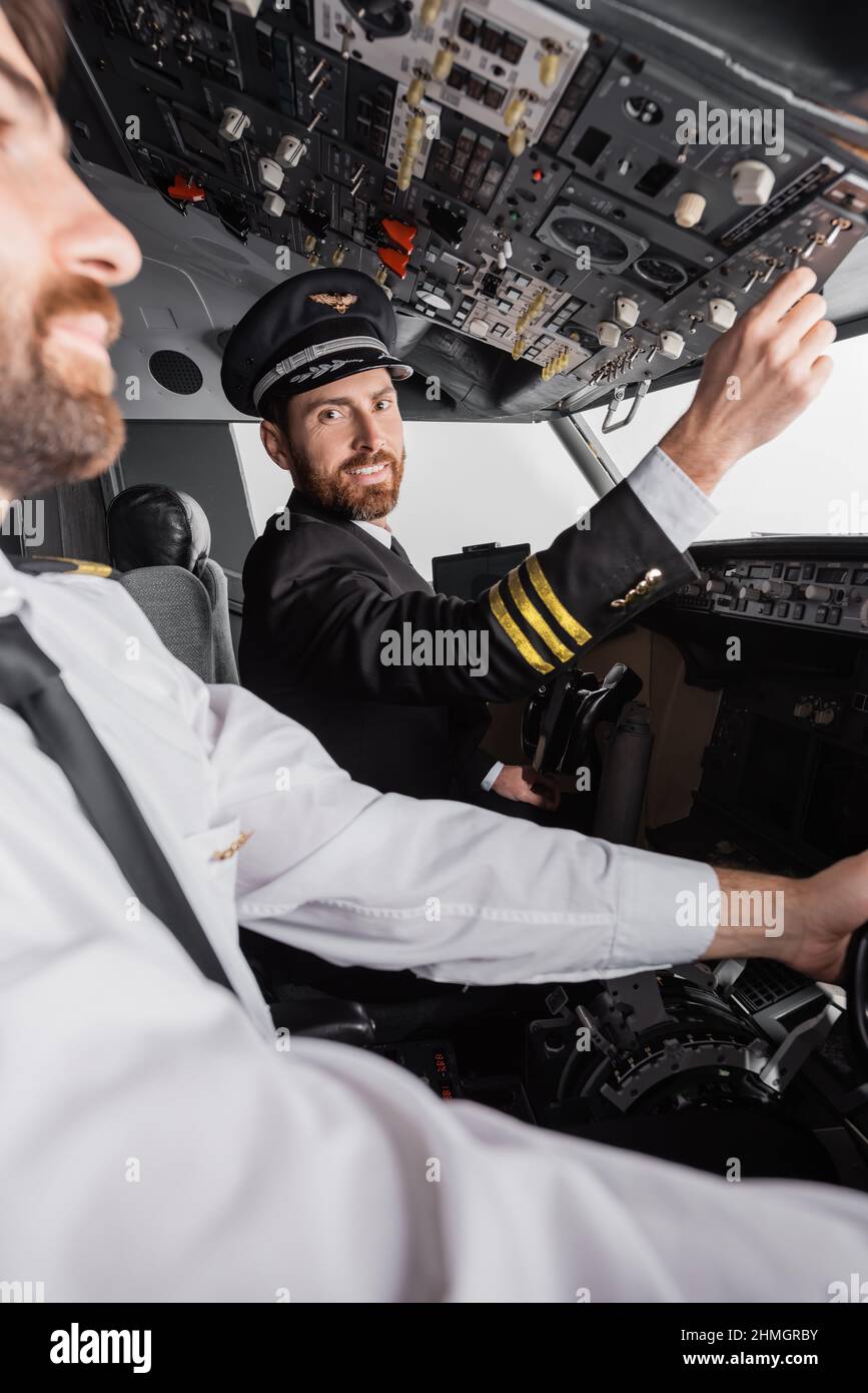 cheerful captain in cap and uniform reaching overhead panel near co ...
