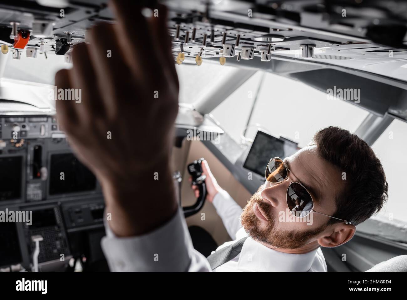 high angle view of bearded pilot in sunglasses reaching overhead panel ...