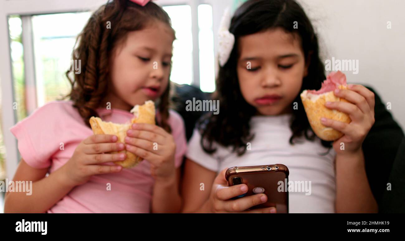 Two little girls hypnotized by cellphone while eating sandwich snack ...