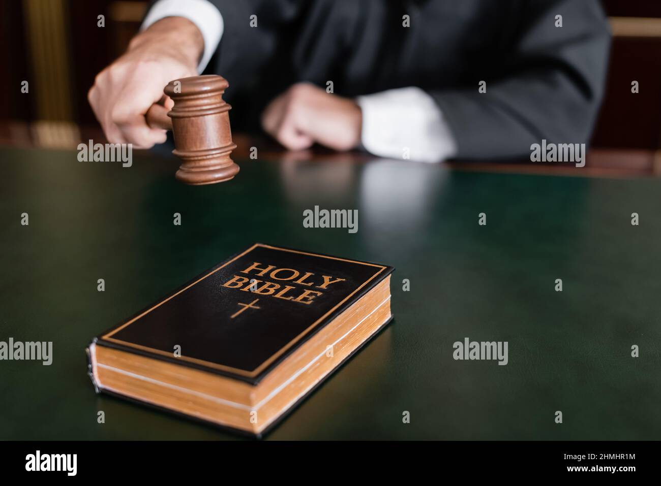 partial view of blurred judge holding gavel near holy bible on desk ...