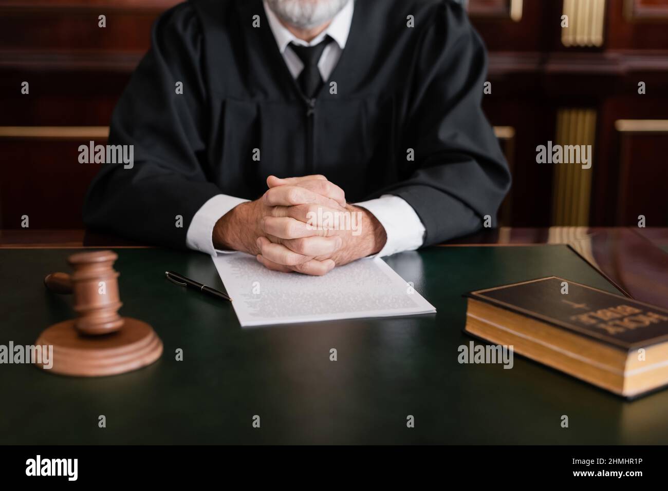 partial view of judge sitting with clenched hands near bible, gavel and ...