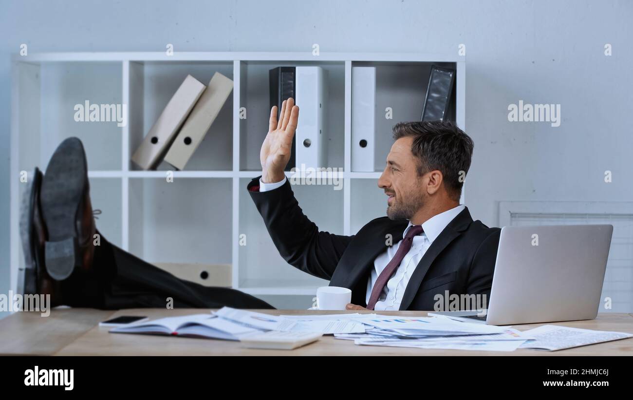 side view of happy man waving hand while sitting with legs on desk at ...