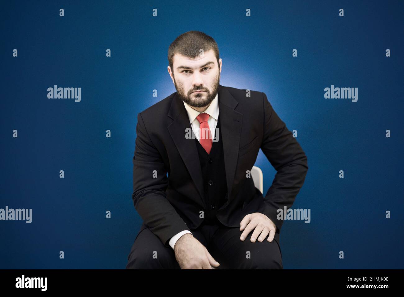 A brutal handsome man with a beard in a suit and red tie looks ahead ...
