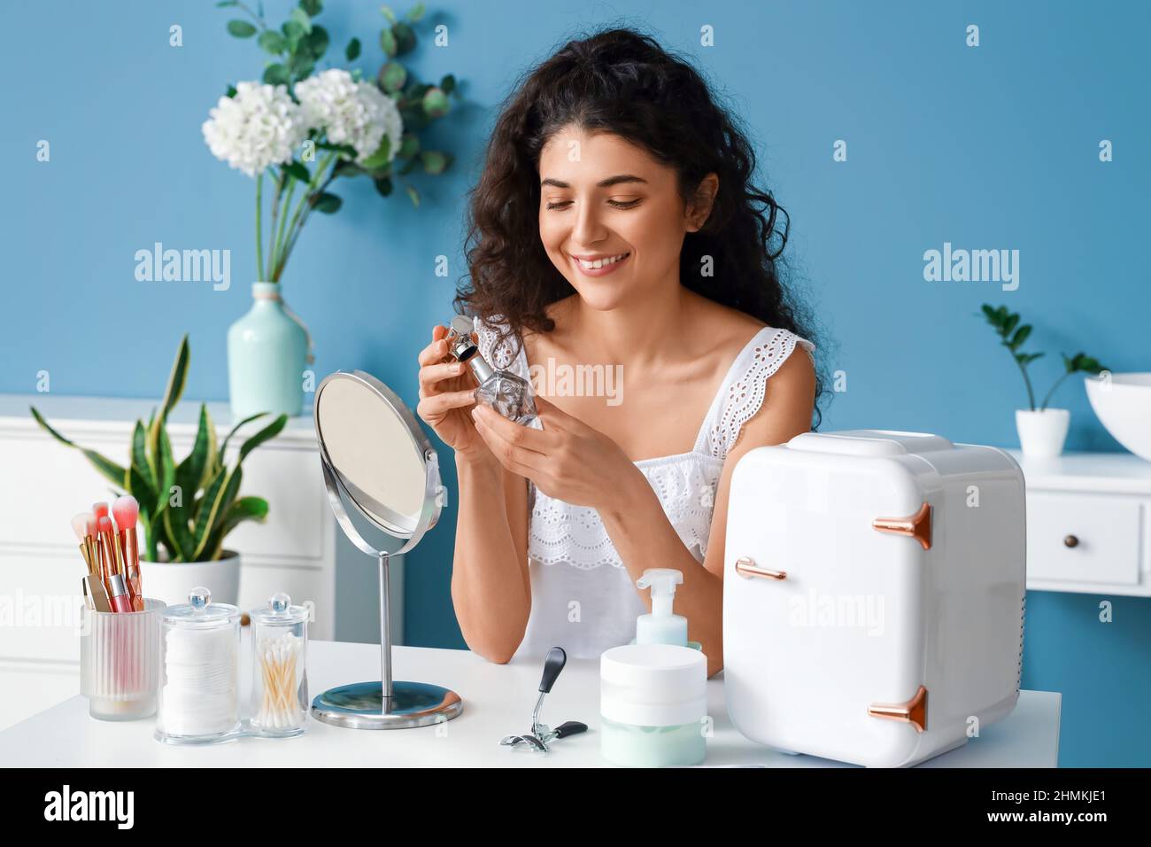 Beautiful woman holding perfume bottle at table with small refrigerator ...