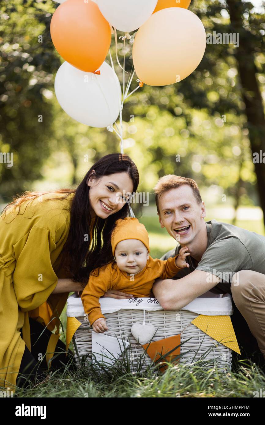 Little baby boy sitting in wicker basket with colorful balloons while ...
