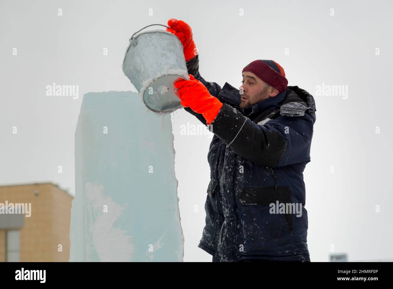 An ice town builder in a blue hooded jacket pours water from a bucket ...
