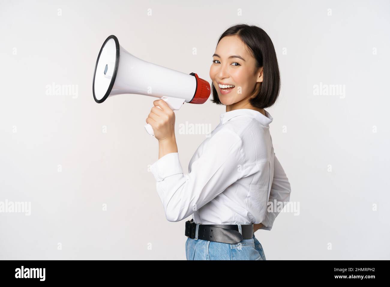 Beautiful young asian woman talking in megaphone, screams in speakerphone and smiling, making ...