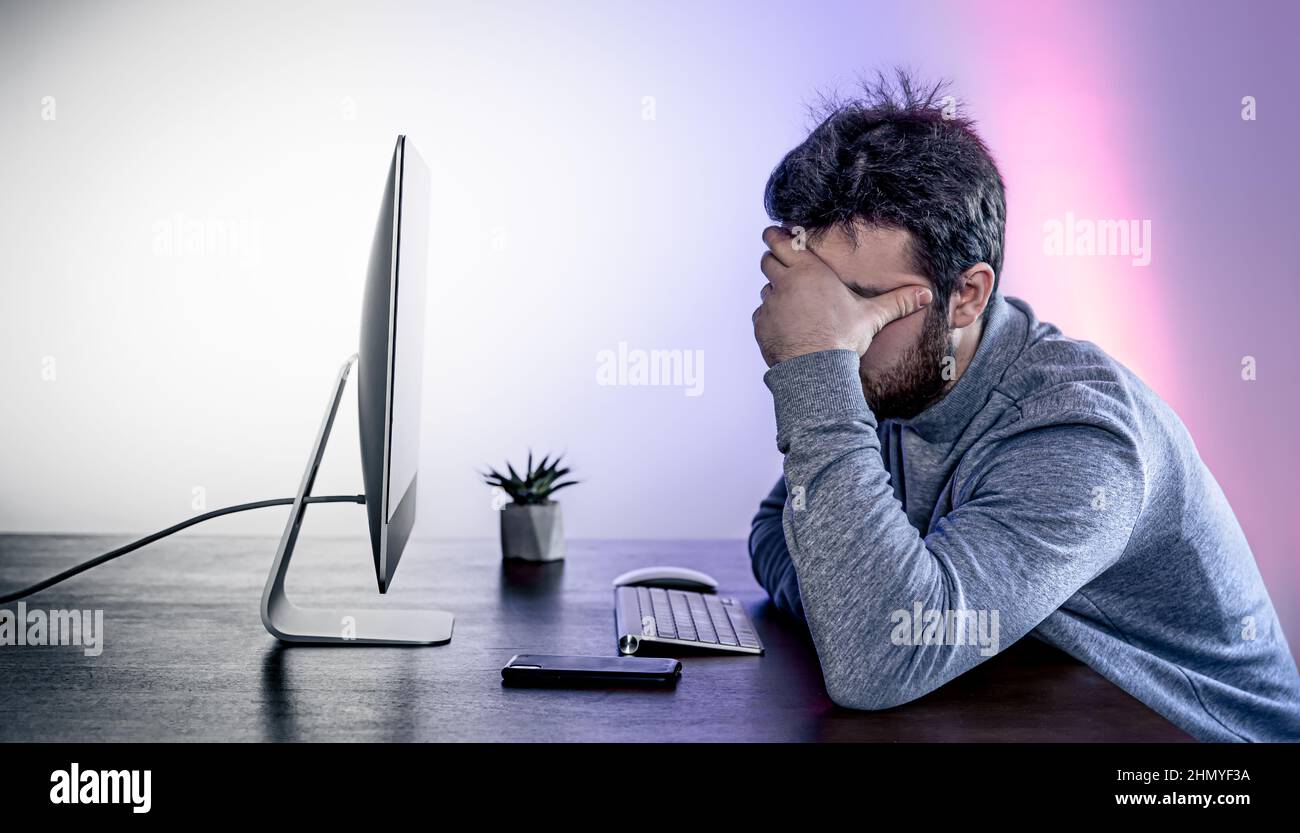 A tired man sits in front of a computer, covering his face with his ...