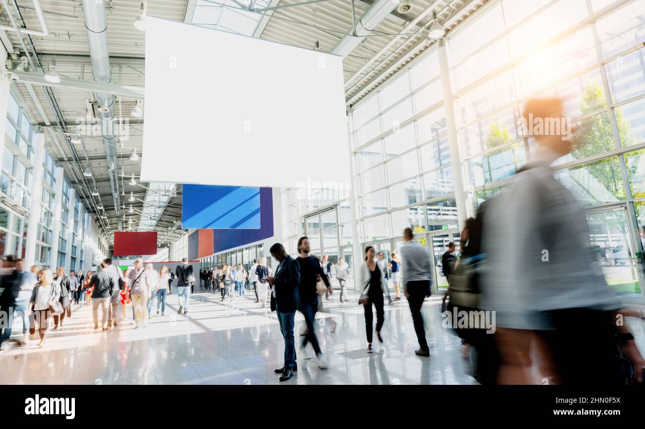 Many trade fair visitors walk under a blank billboard through a hall ...