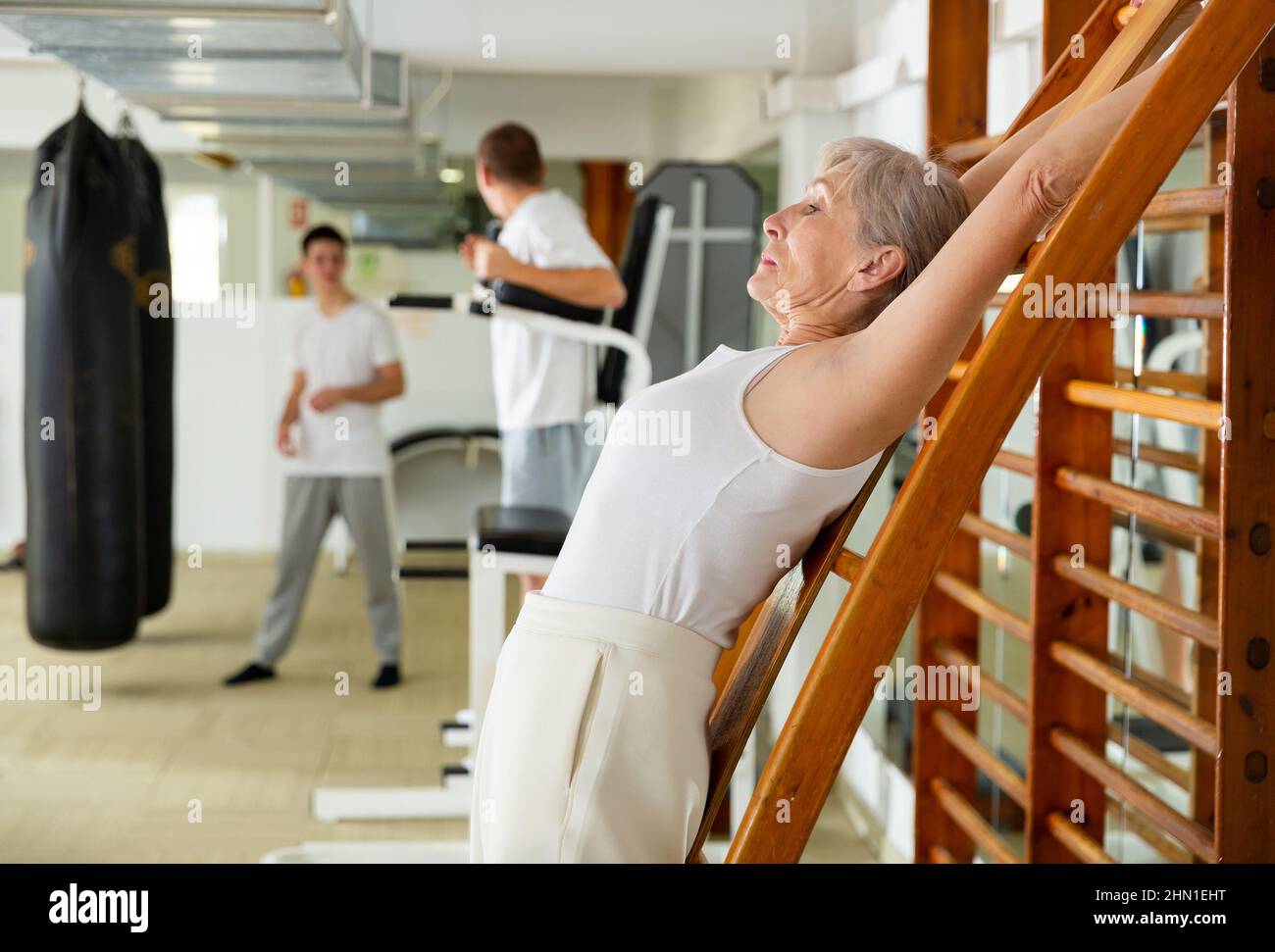 Elderly woman doing back stretching exercise on wall bar in gym Stock ...