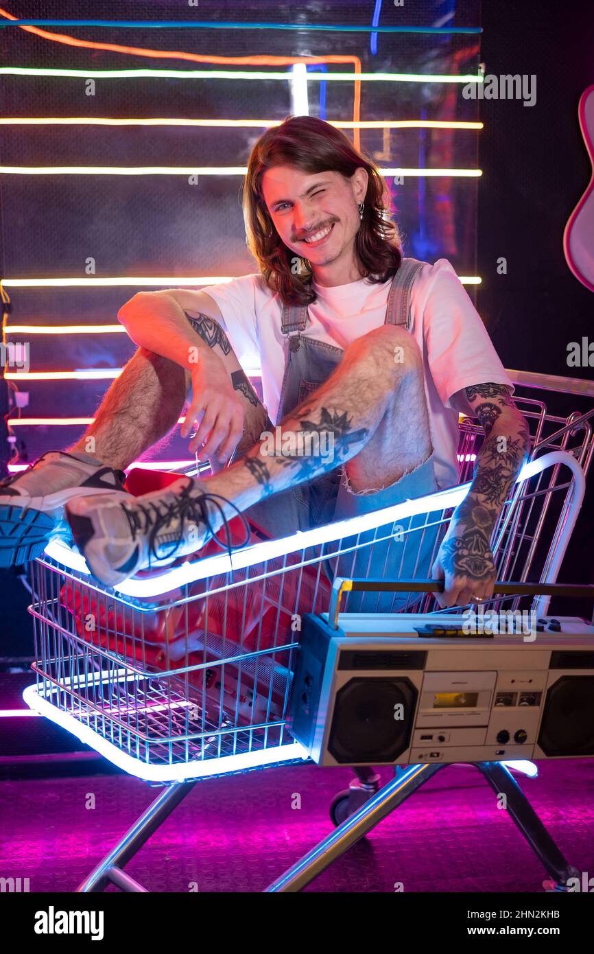 Millennial man sitting in a shopping cart in old retro store buy a tape ...