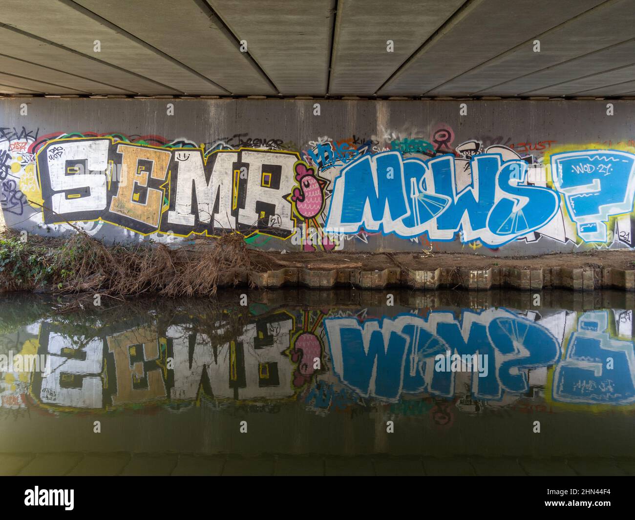 Colourful graffiti under a road bridge over a canal, with reflections ...