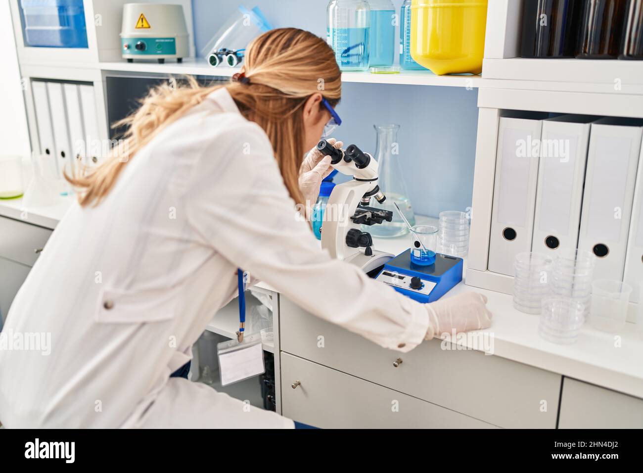 Young blonde woman wearing scientist uniform using microscope at ...