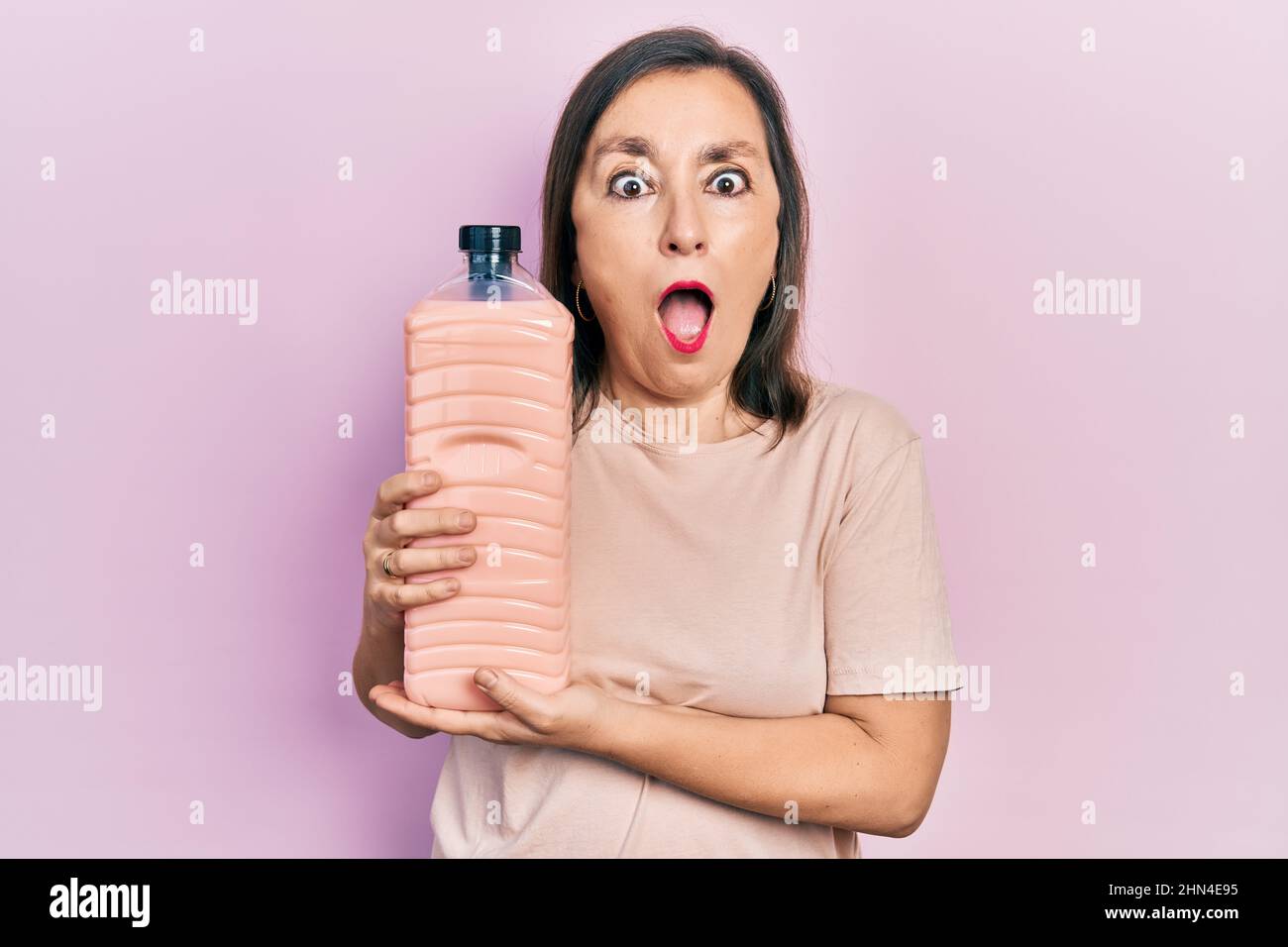 Middle age hispanic woman holding detergent bottle afraid and shocked with surprise and amazed ...