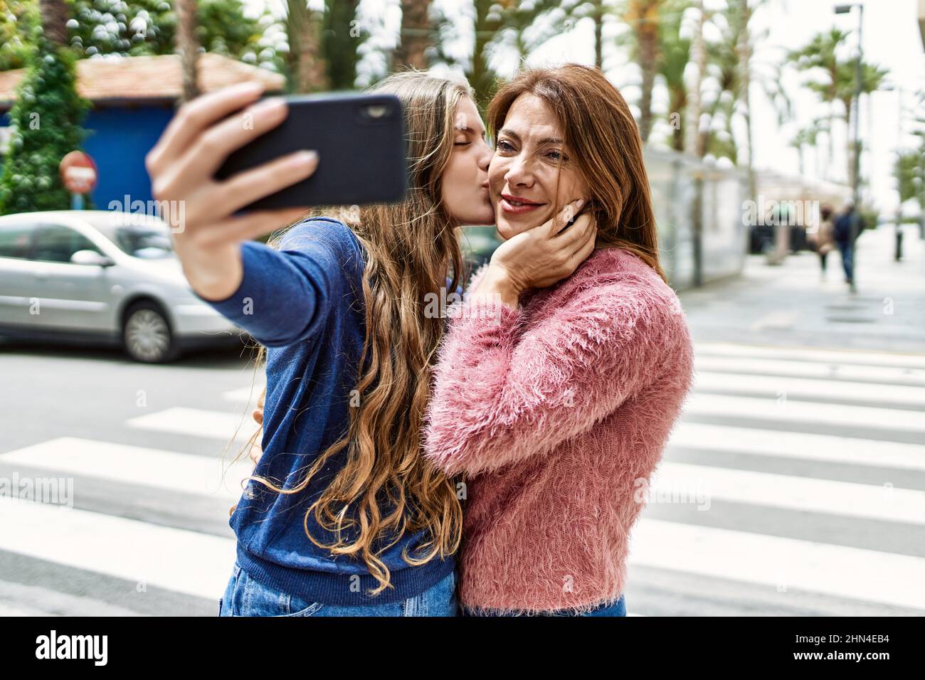 Mother and daughter hugging each other and kissing making selfie by the smartphone at street ...