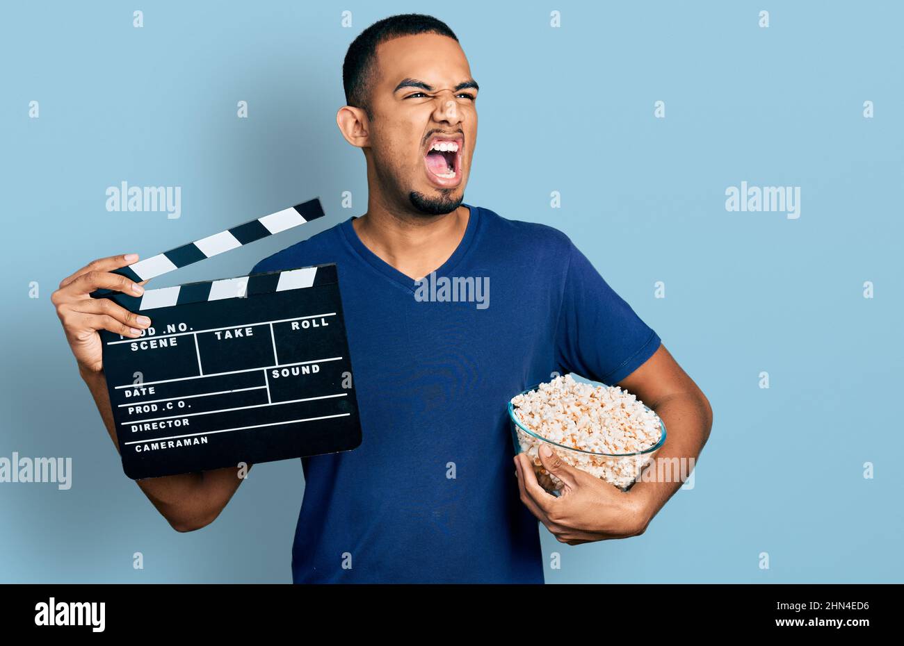 Young african american man eating popcorn holding film clapboard angry ...