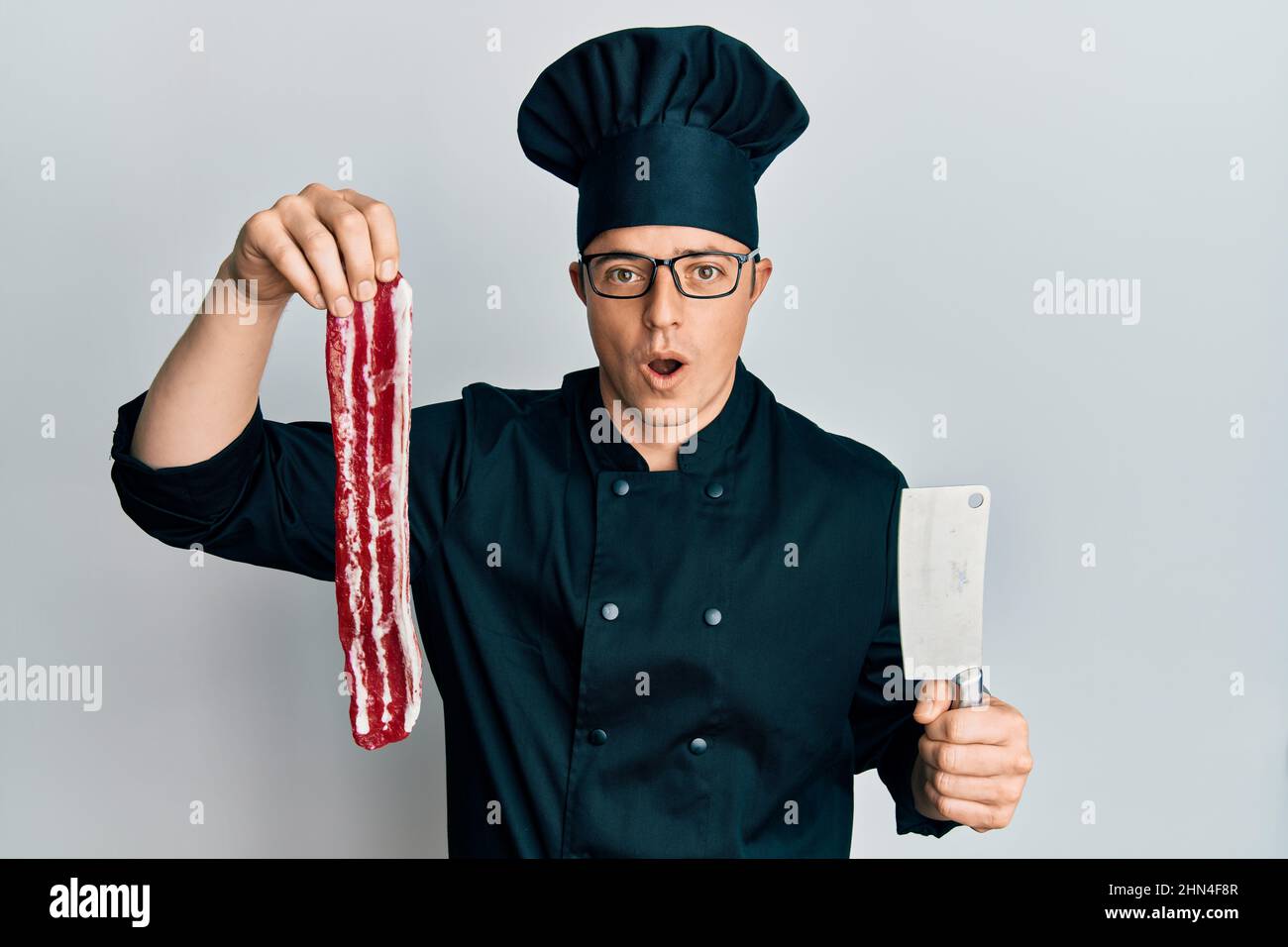 Handsome young man wearing chef uniform holding bacon and knife afraid ...