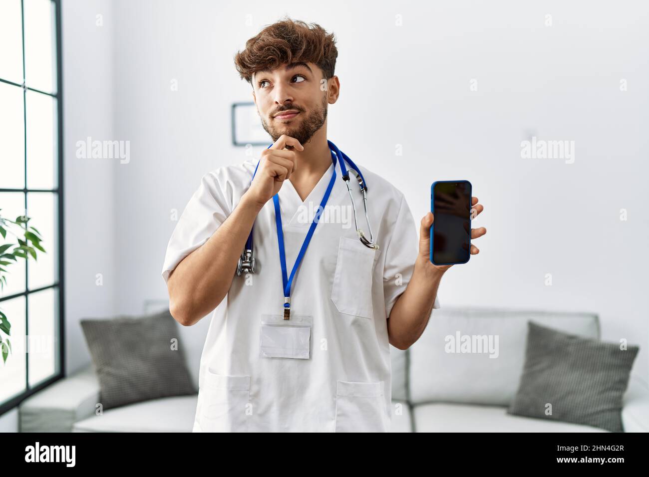 Young arab man wearing doctor uniform and stethoscope holding smartphone serious face thinking ...