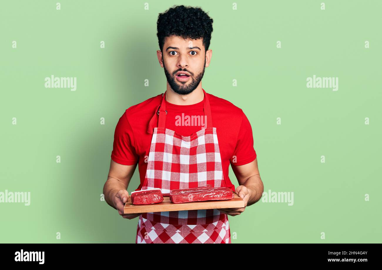 Young arab man with beard holding board with raw meat afraid and ...
