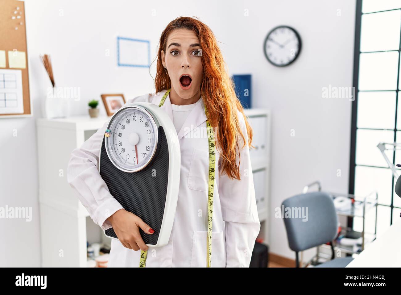 Young redhead woman nutritionist doctor holding weighing machine scared ...