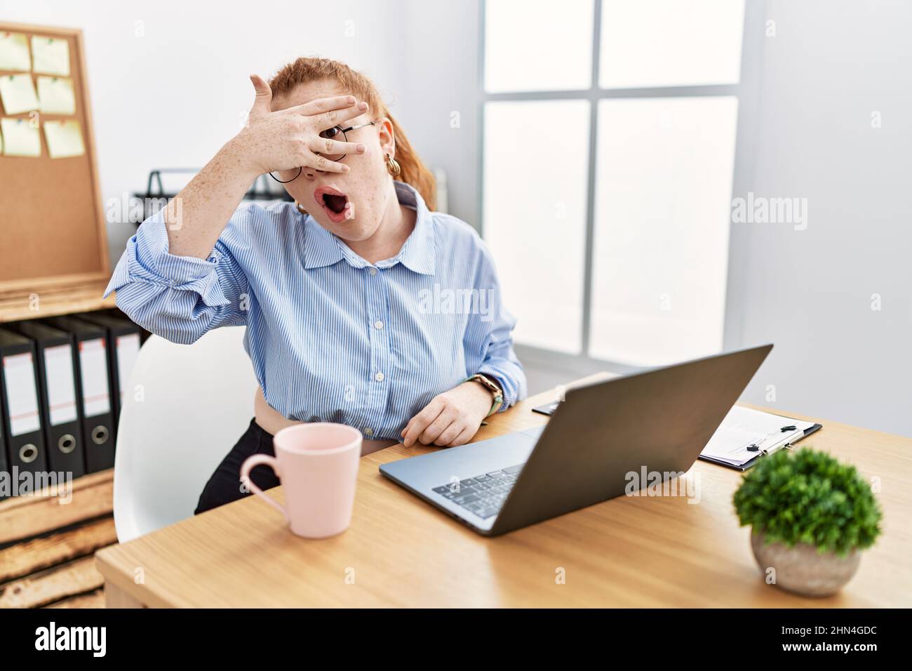 Young redhead woman working at the office using computer laptop peeking ...