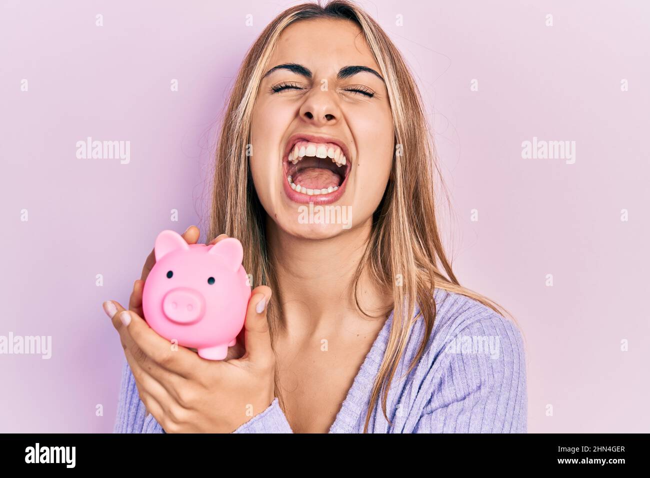Beautiful hispanic woman holding piggy bank angry and mad screaming ...