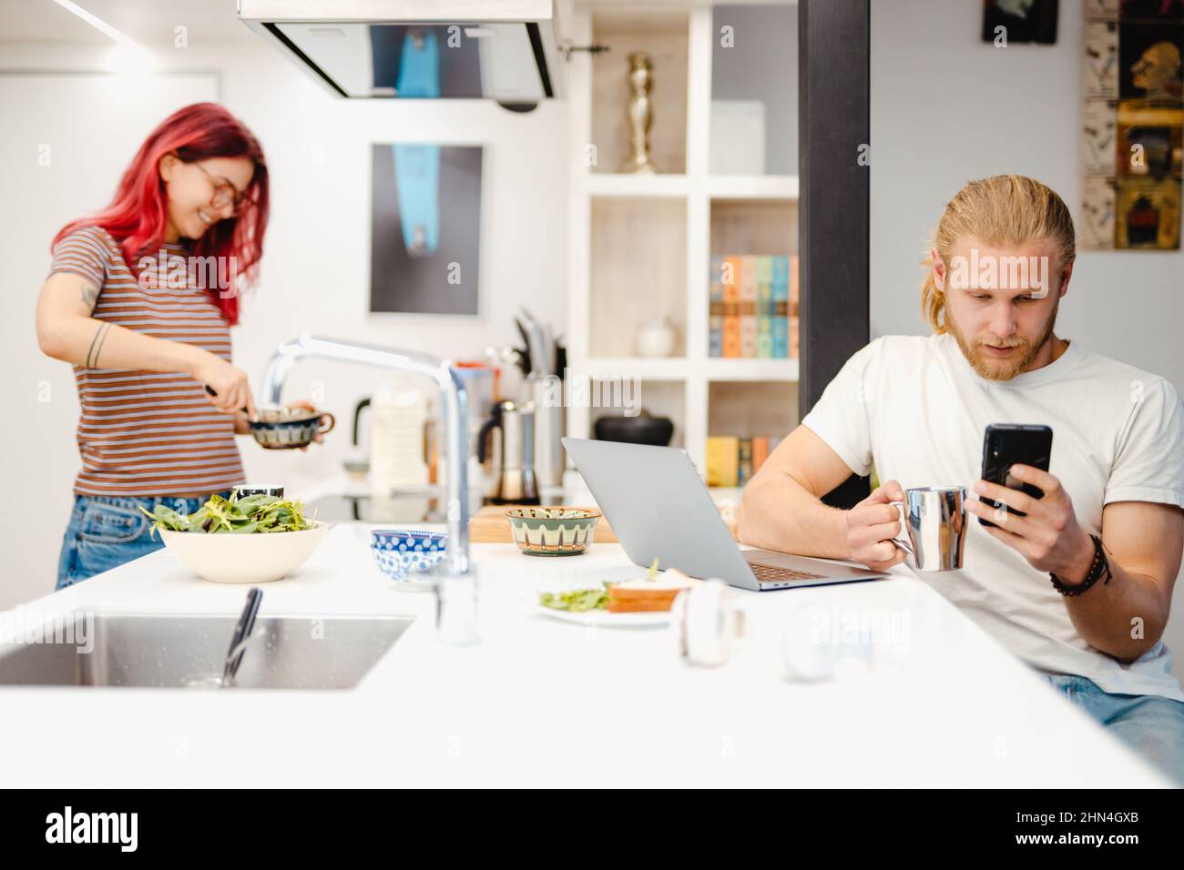 Bearded man using cellphone while his girlfriend cooking in kitchen at ...
