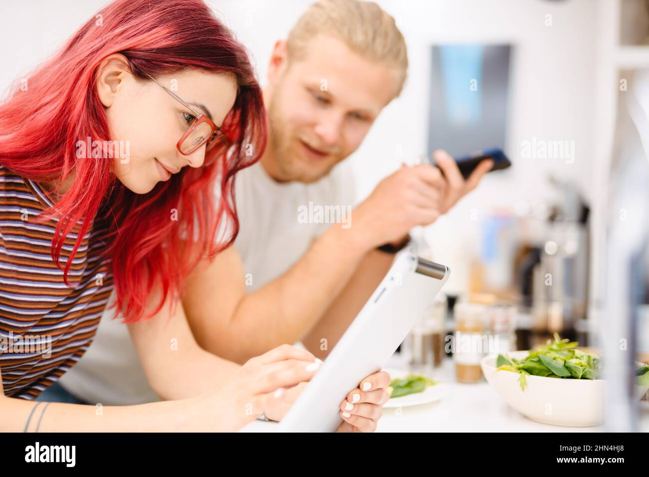 Young white couple using gadgets while having lunch in kitchen at home ...