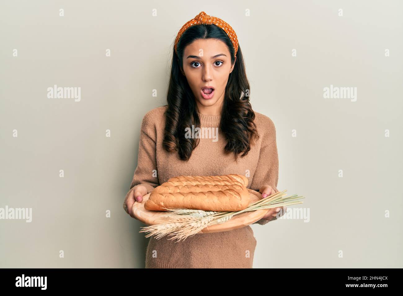 Young hispanic woman holding bread in shock face, looking skeptical and sarcastic, surprised ...