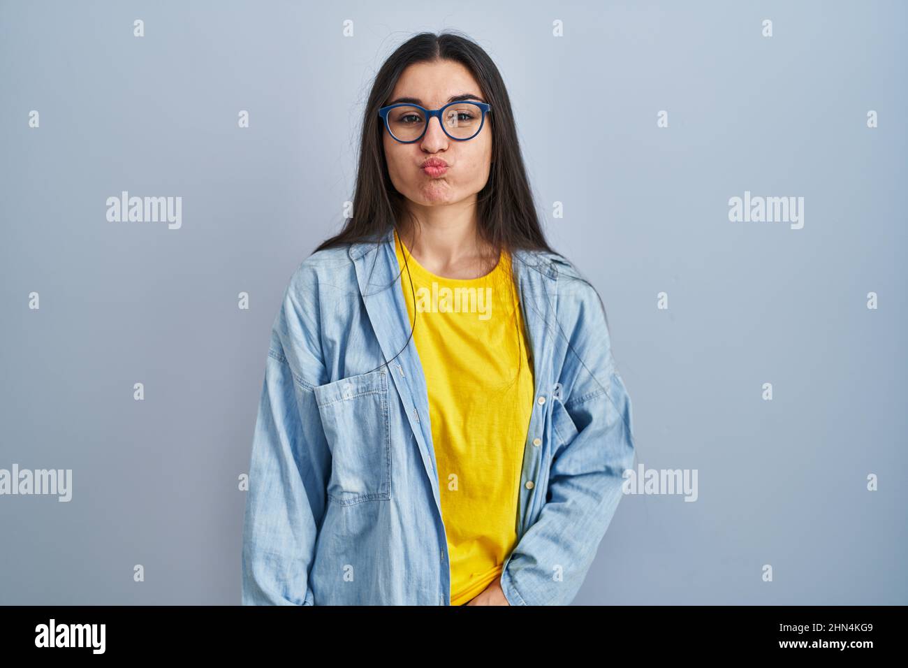 Young hispanic woman standing over blue background puffing cheeks with ...